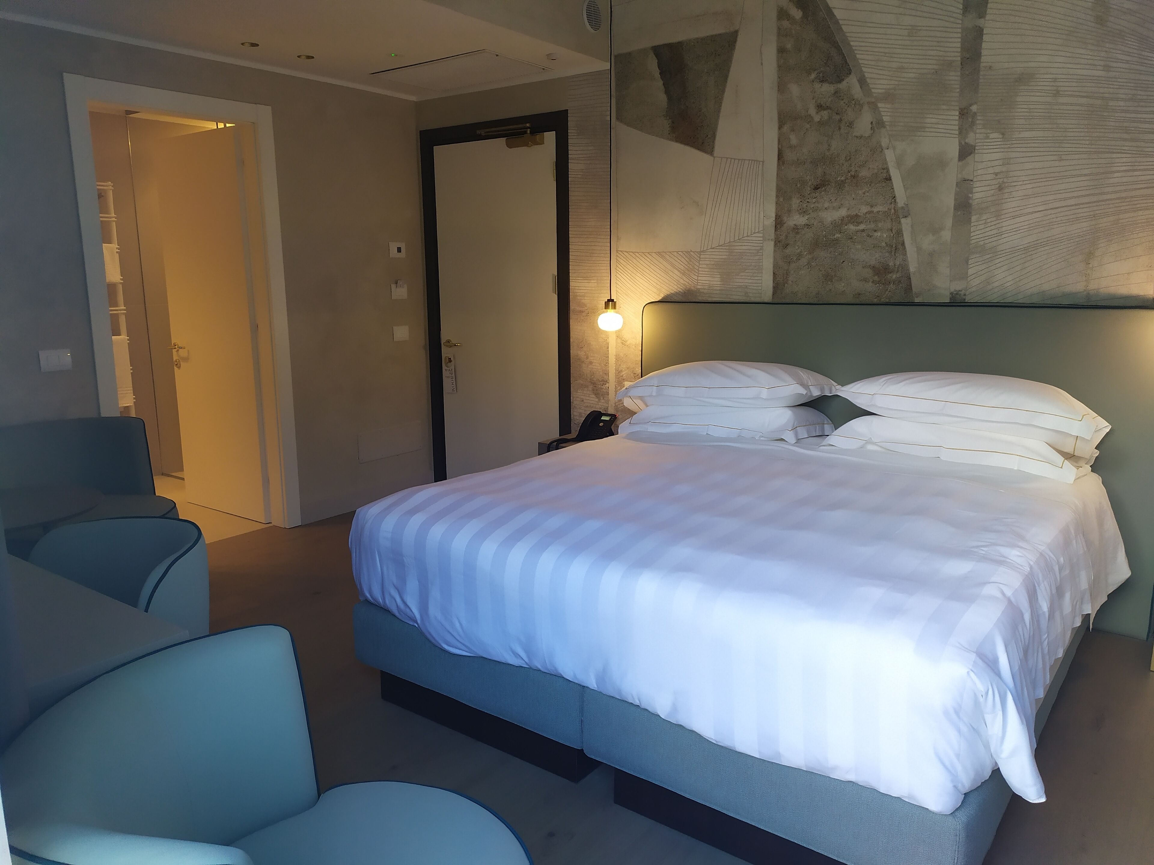 Quarto de hotel com cama, uma cadeira, mesa de cabeceira e luminária fixada na parede.