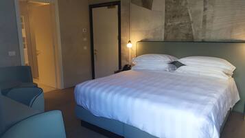 Quarto de hotel com cama, uma cadeira, mesa de cabeceira e luminária fixada na parede.