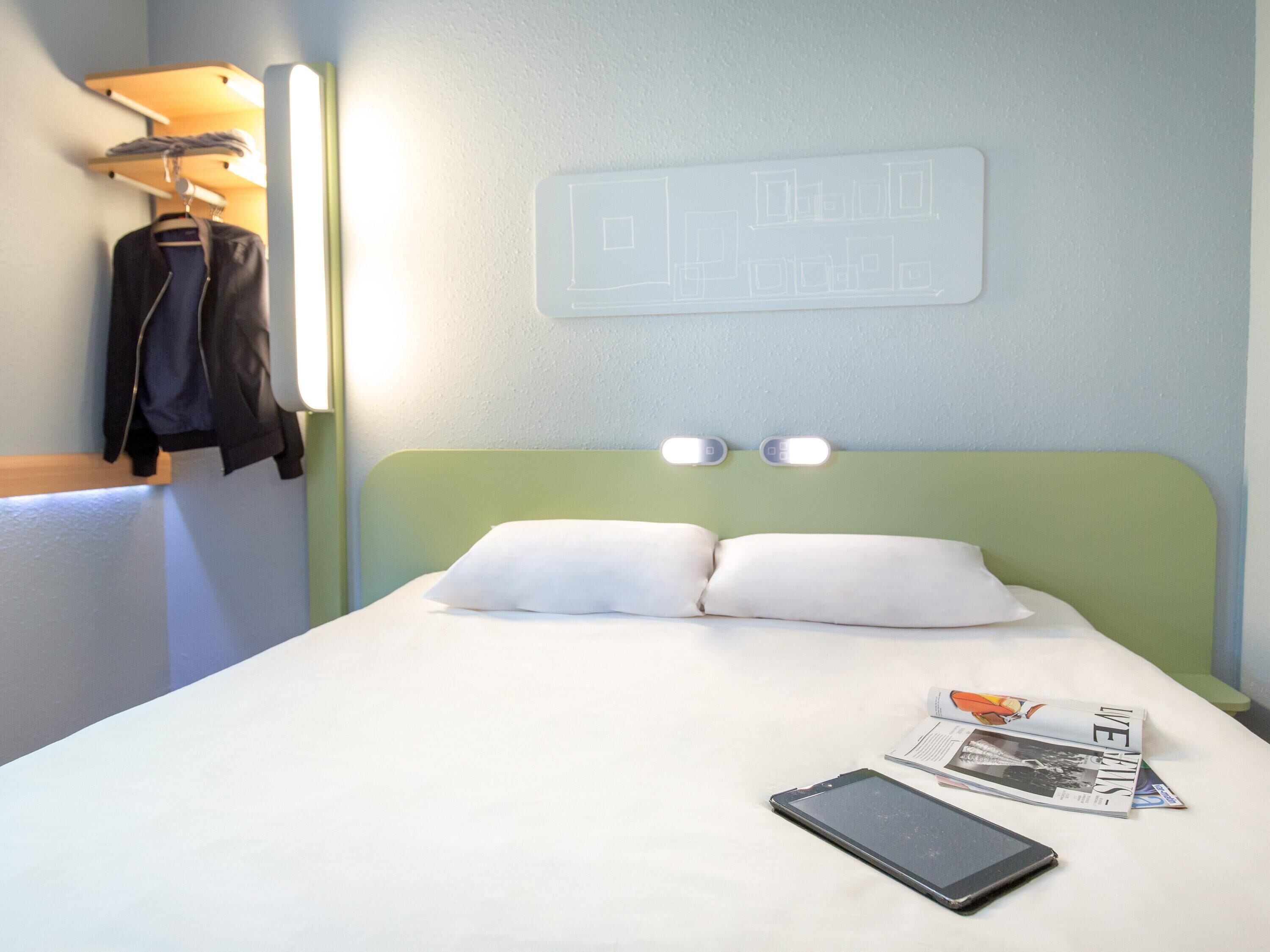 Uma cama bem arrumada com almofadas brancas e cabeceira verde, uma mesa de cabeceira com um tablet e uma revista, e um luminária fixada na parede.