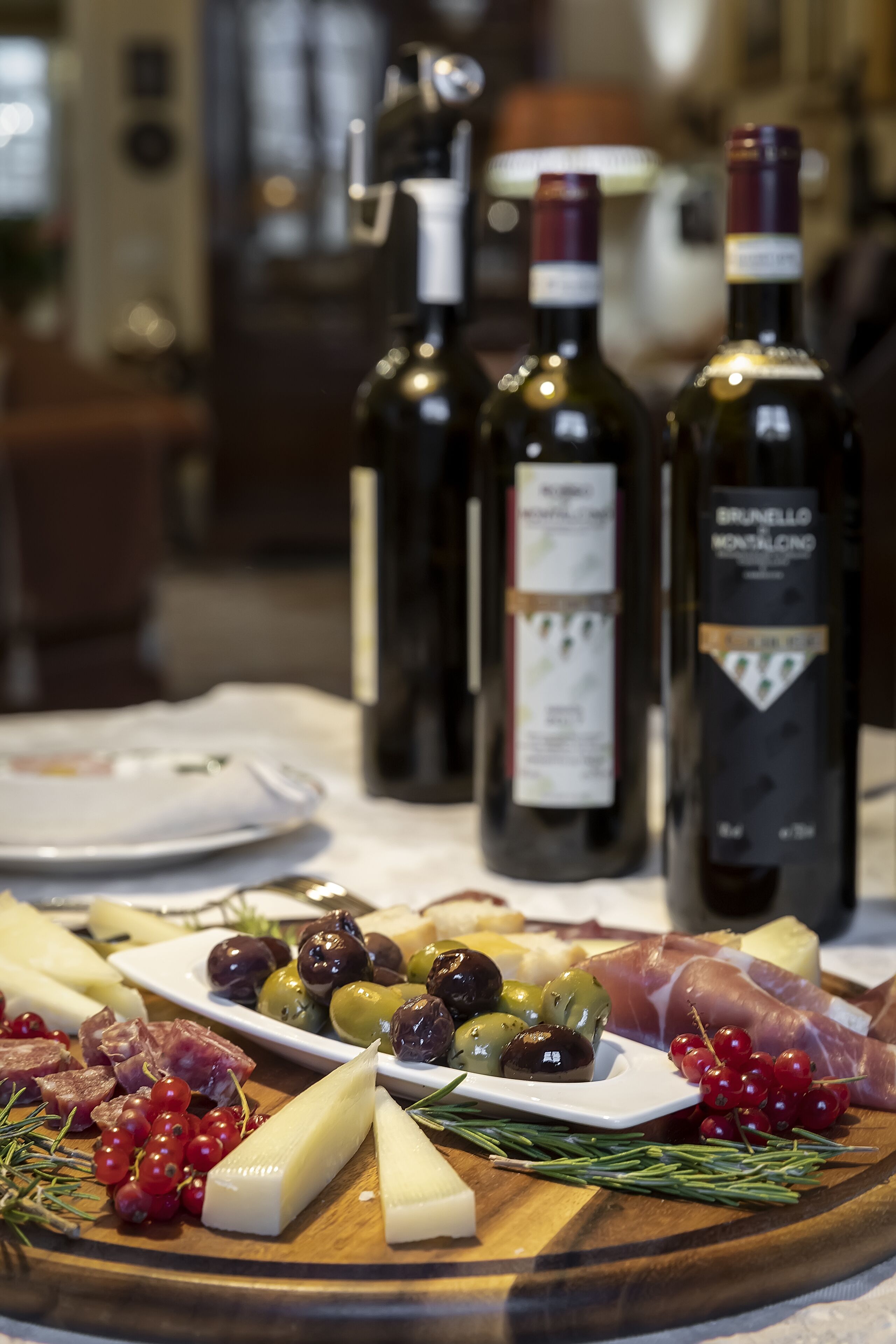 Uma tábua de frios com diversos queijos, carnes e frutas, acompanhada por garrafas de vinho.