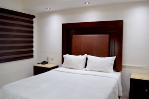 Quarto de hotel com cama grande, cabeceira de madeira, mesa de cabeceira com telefone e janela com persianas.