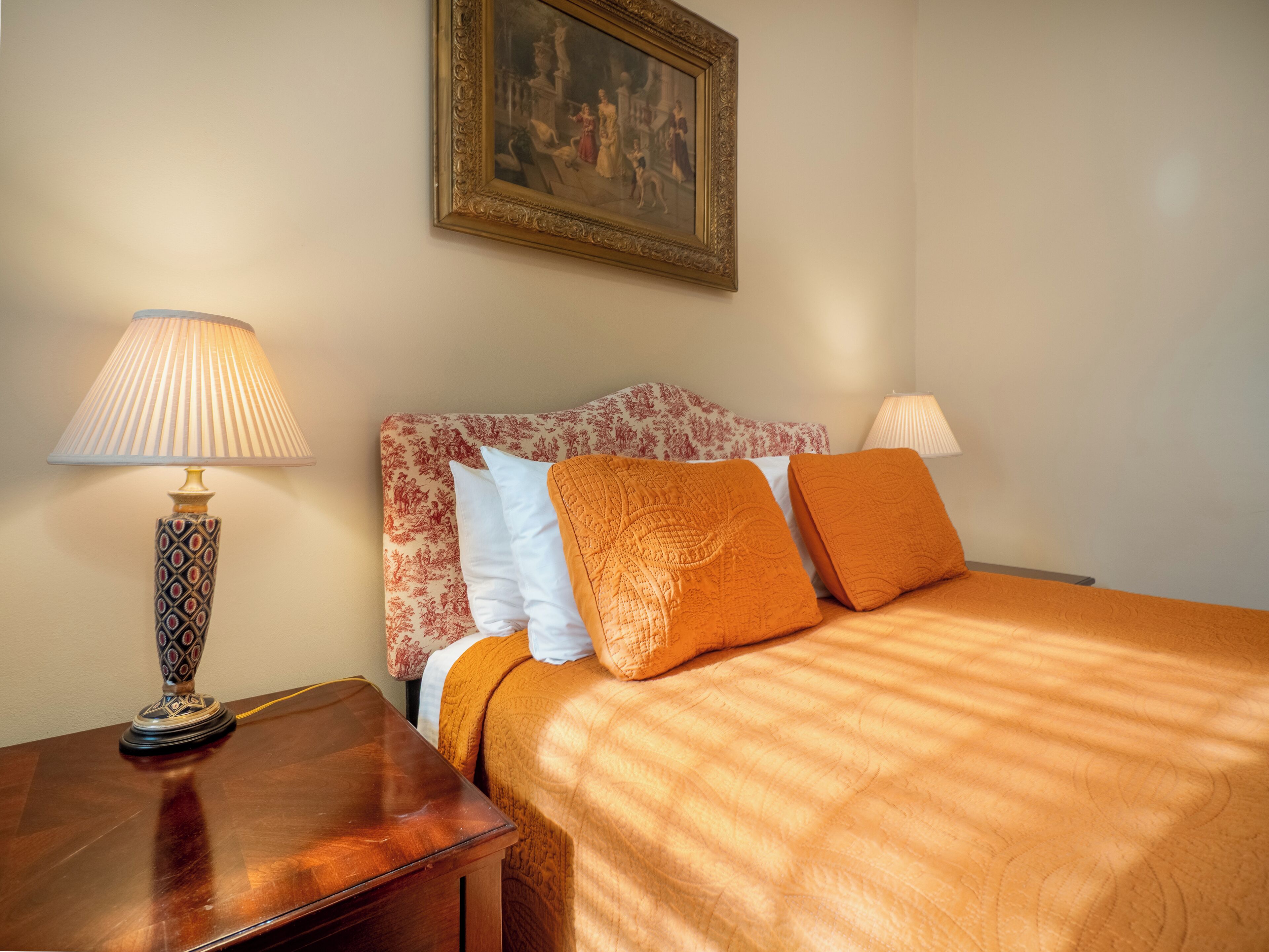 Cama bem arrumada com cabeceira estampada, duas luminárias laterais e um quadro na parede.