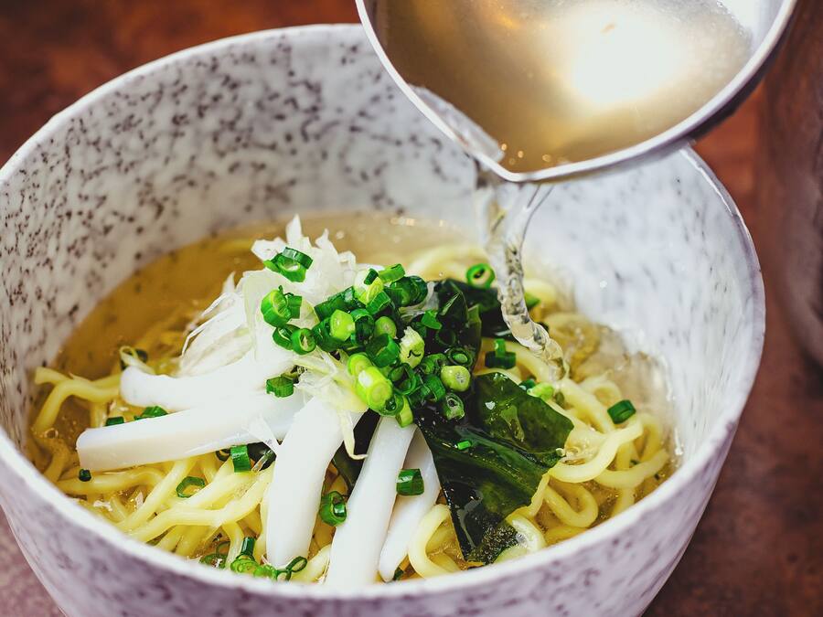 Uma tigela de ramen com macarrão, cebolinha e alga marinha sendo adicionada.