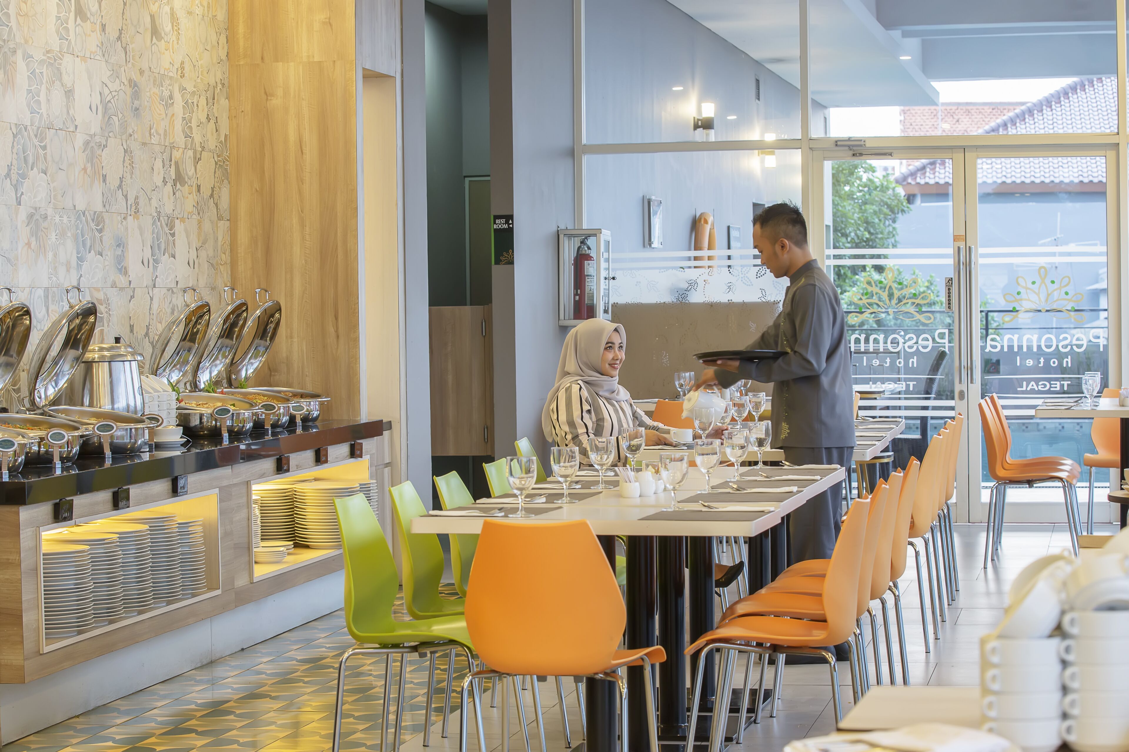 Restaurante com buffet, garçom atendendo um cliente e vista para o exterior através de grandes janelas.
