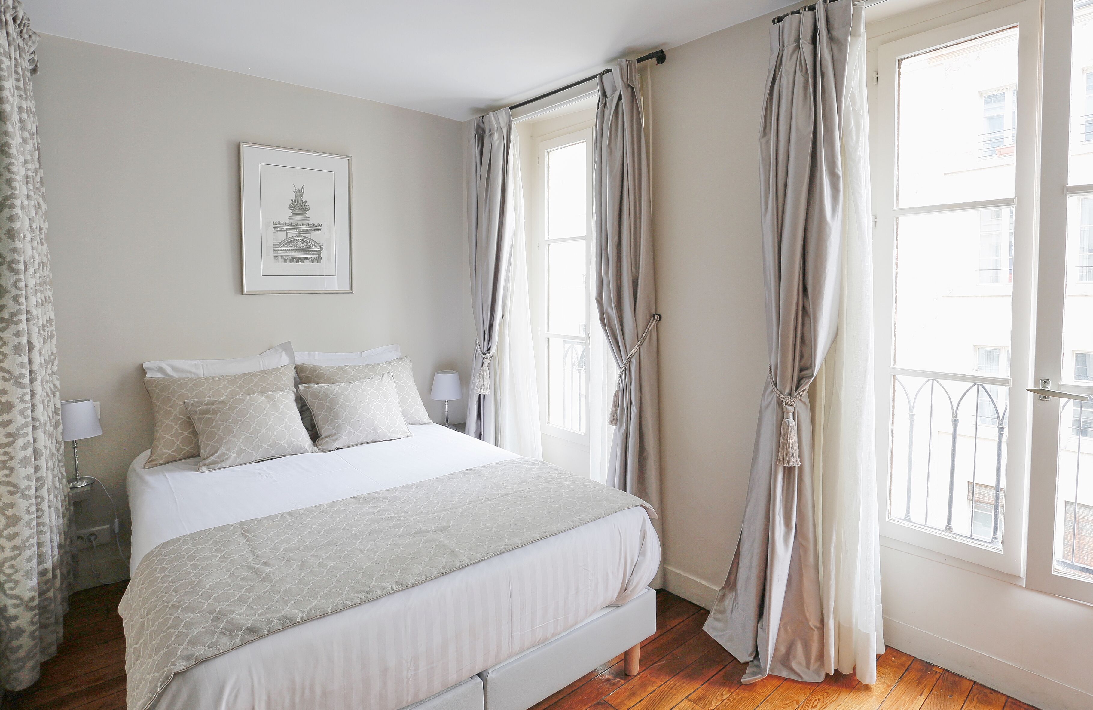 Quarto com cama, cortinas, mesa de cabeceira, luminária e um quadro na parede.