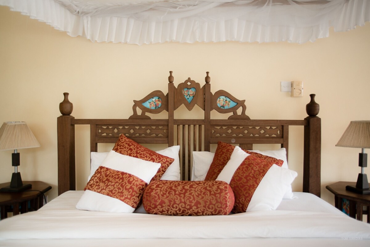 Cama bem arrumada com almofadas estampadas, cabeceira de madeira com detalhes decorativos e duas luminárias de cabeceira.