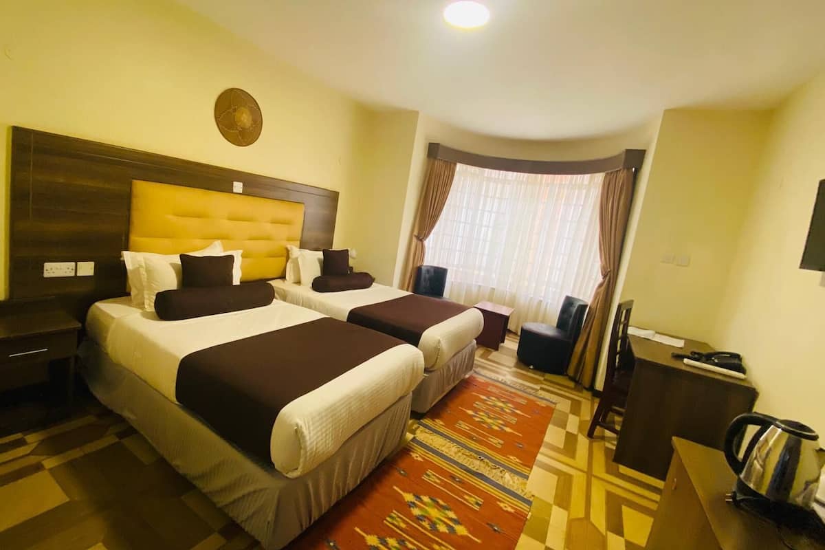 Quarto de hotel com duas camas, uma cabeceira grande, um relógio, uma escrivaninha com chaleira e uma janela com cortinas.