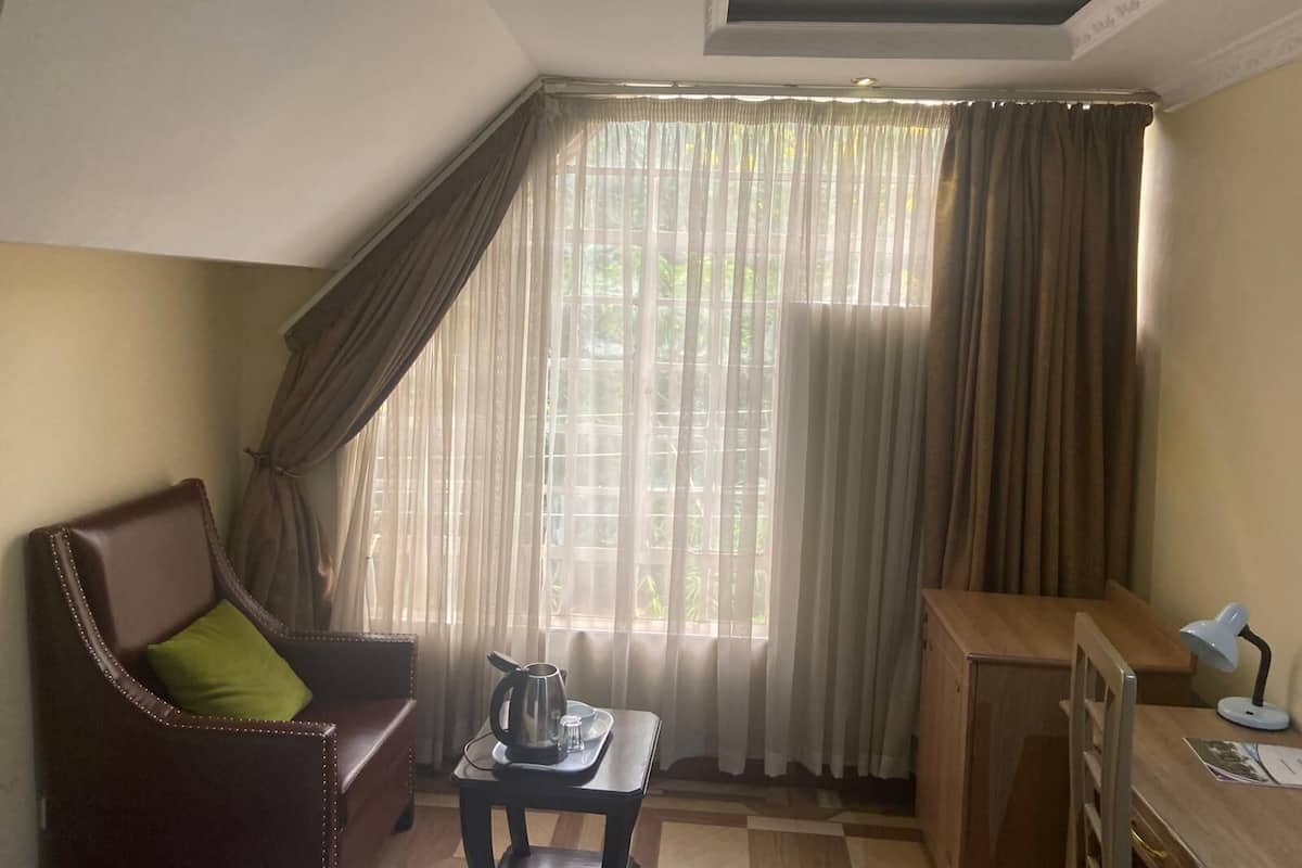 Quarto com poltrona de couro marrom, mesa pequena com chaleira e bandeja, escrivaninha com luminária e janela com cortinas leves.