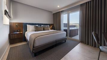 Quarto de hotel moderno com uma cama grande, uma mesa de cabeceira, uma cadeira e vista para a cidade através de uma janela ampla.