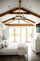 Quarto com cama grande, vigas de madeira no teto, lustre, cômoda e vista para o verde lá fora.