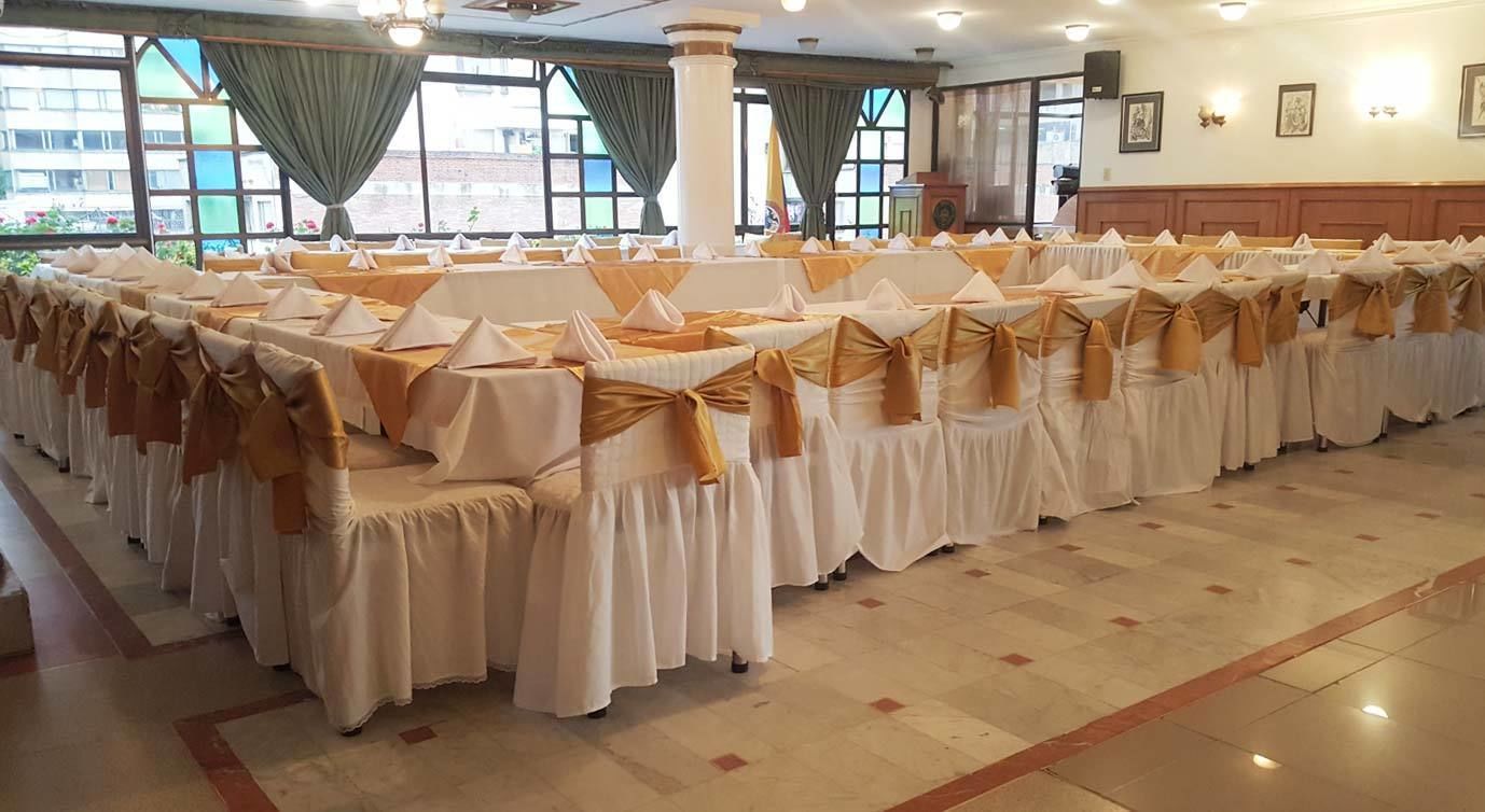 Uma sala de banquetes com fileiras de mesas cobertas por toalhas brancas e fitas laranja, grandes janelas com cortinas e piso ladrilhado.