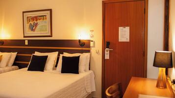 Quarto de hotel com uma cama grande, uma mesa de trabalho e uma cadeira. Há uma pintura na parede e uma porta com um aviso.
