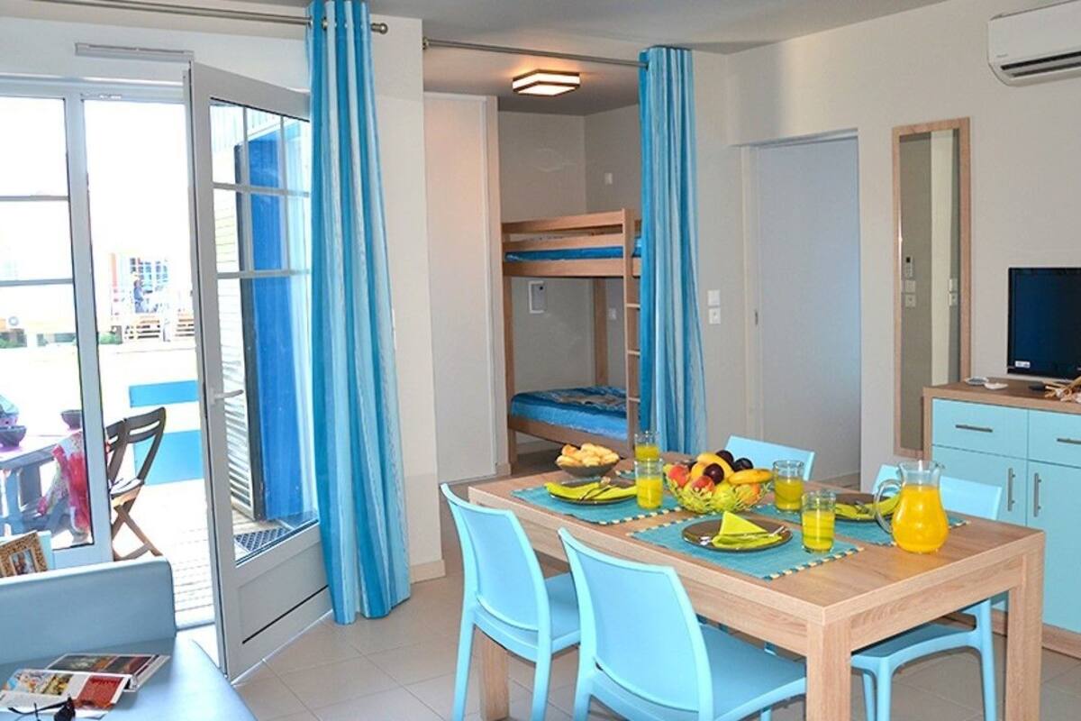 Quarto com mesa de jantar posta para dois, uma cama beliche, televisão e janela com cortinas.