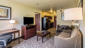 Quarto de hotel com secretária, cadeira, sofá, televisão, frigorífico e kitchenette.
