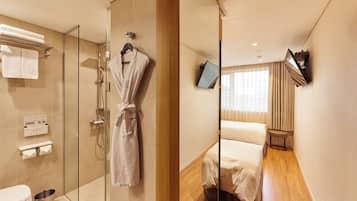 Quarto de hotel com chuveiro de vidro, cama sobre piso de madeira e televisão montada na parede.