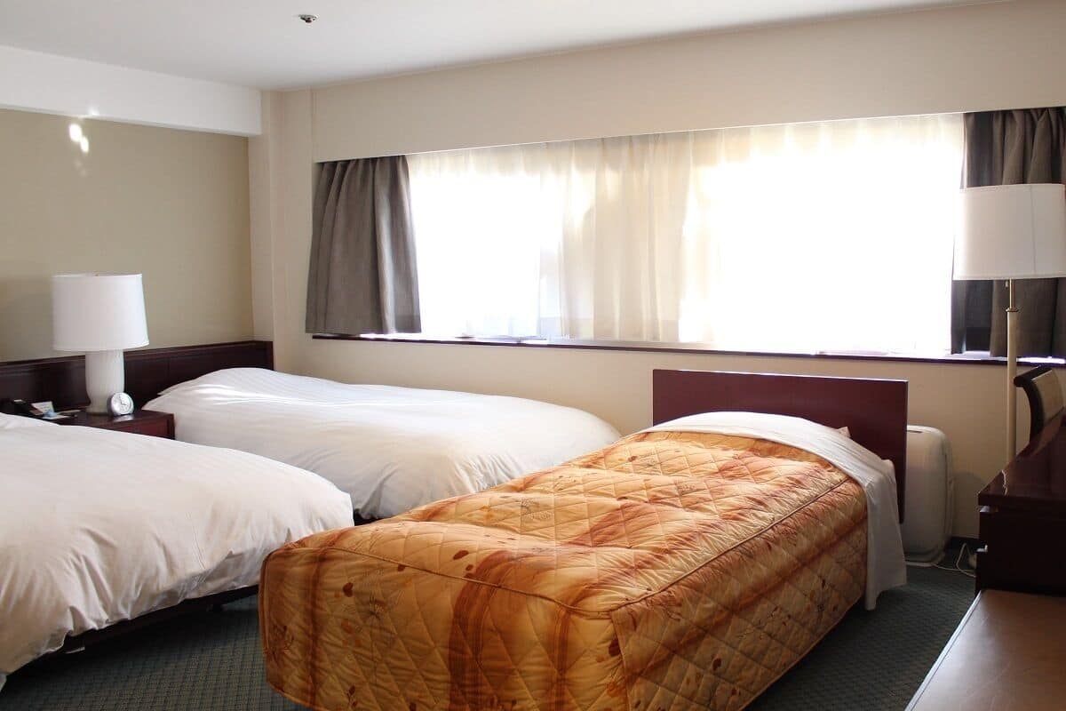 Quarto de hotel com duas camas de solteiro, uma escrivaninha, uma cadeira, um abajur e uma janela com cortinas.