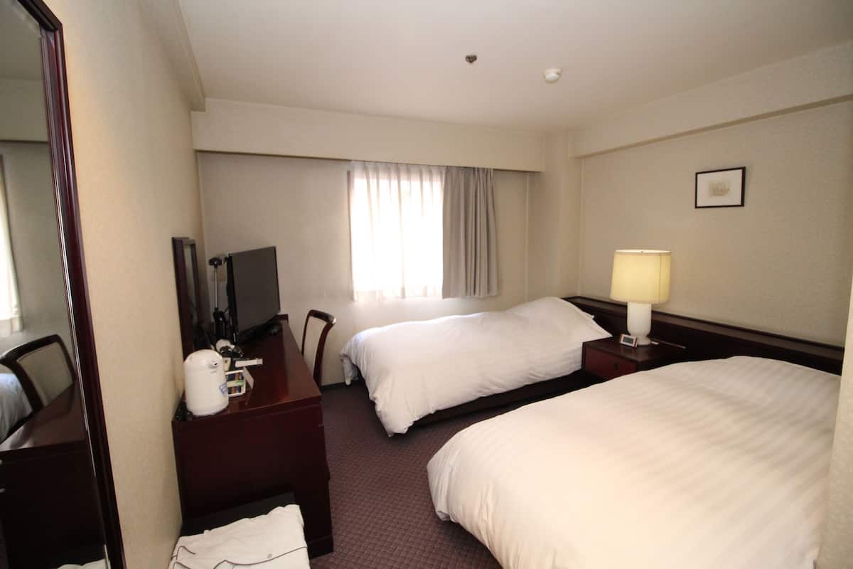 Quarto de hotel com duas camas, uma escrivaninha com cadeira, luminária de mesa, espelho e janela com cortinas.