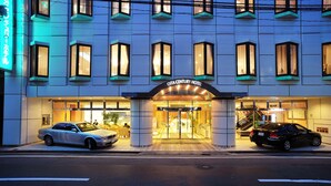 Entrada do Hotel Oita Century com dois carros estacionados em frente.