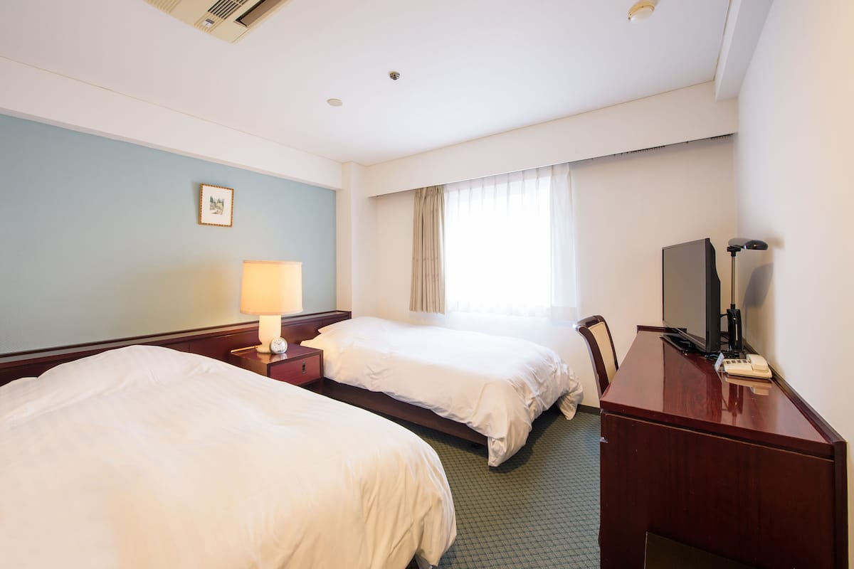 Quarto de hotel com duas camas, uma escrivaninha com luminária, televisão e janela com cortinas.