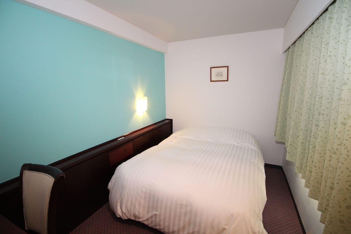 Quarto de hotel com cama, mesa de cabeceira, cadeira, luminária na parede e janela com cortinas.