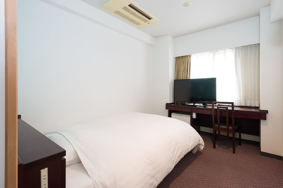 Quarto de hotel com cama, escrivaninha com televisão e janela com cortinas.
