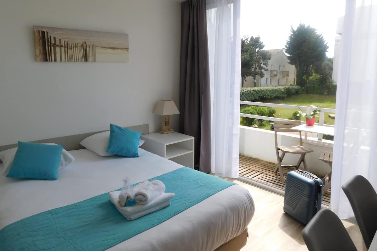 Quarto de hotel com cama, mesa de cabeceira, luminária, janela com cortinas, varanda com cadeira e mesa, e uma mala.
