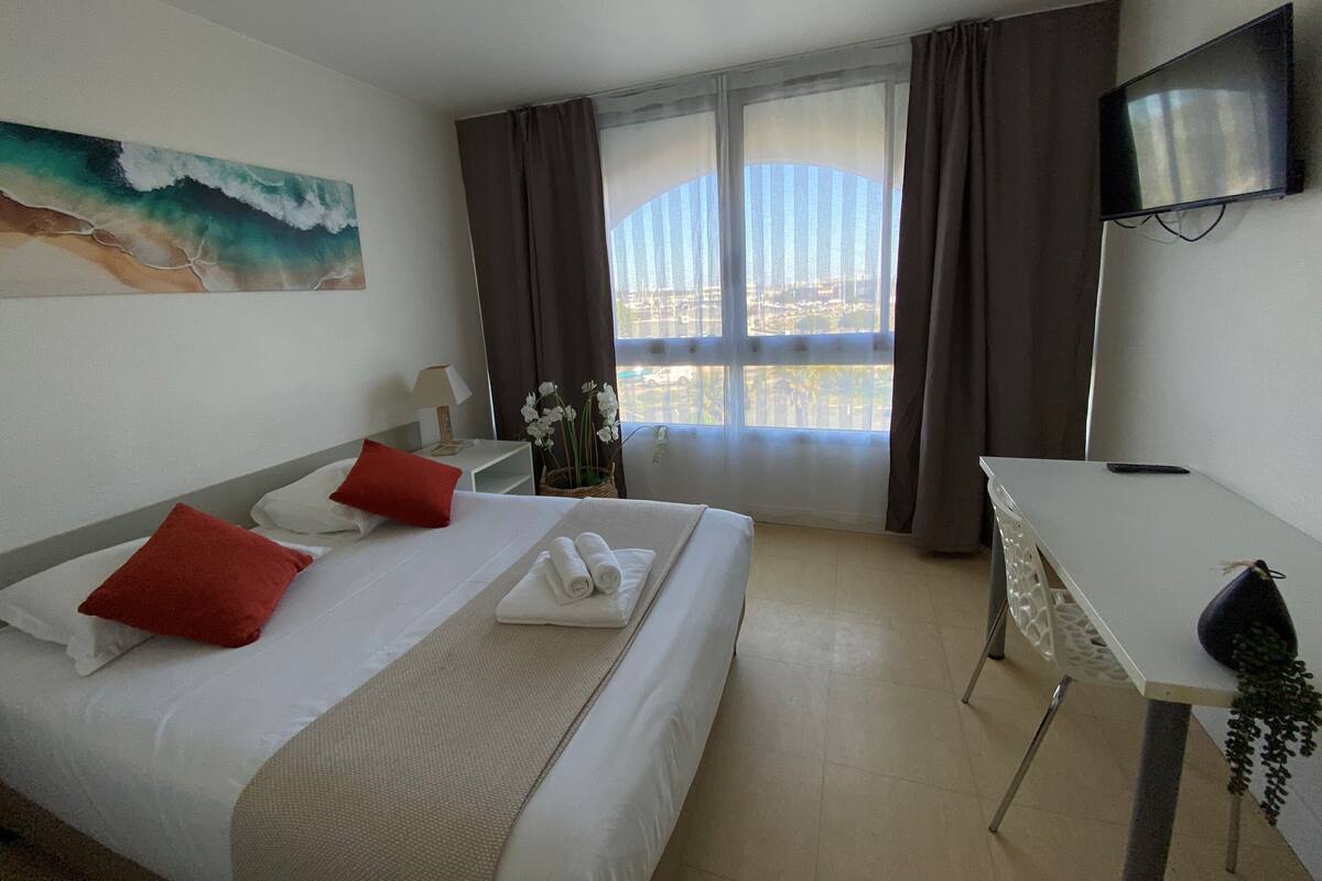 Quarto de hotel com cama, escrivaninha, televisão, janela com cortinas e um quadro na parede.