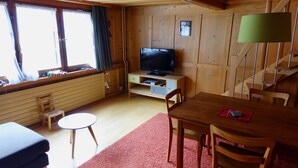 Interior de uma cabana de madeira com uma televisão em um armário de madeira, uma mesa de jantar com cadeiras, uma mesinha redonda e um tapete vermelho.