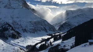 Paisagem montanhosa coberta de neve, com um vale e alguns edifícios.