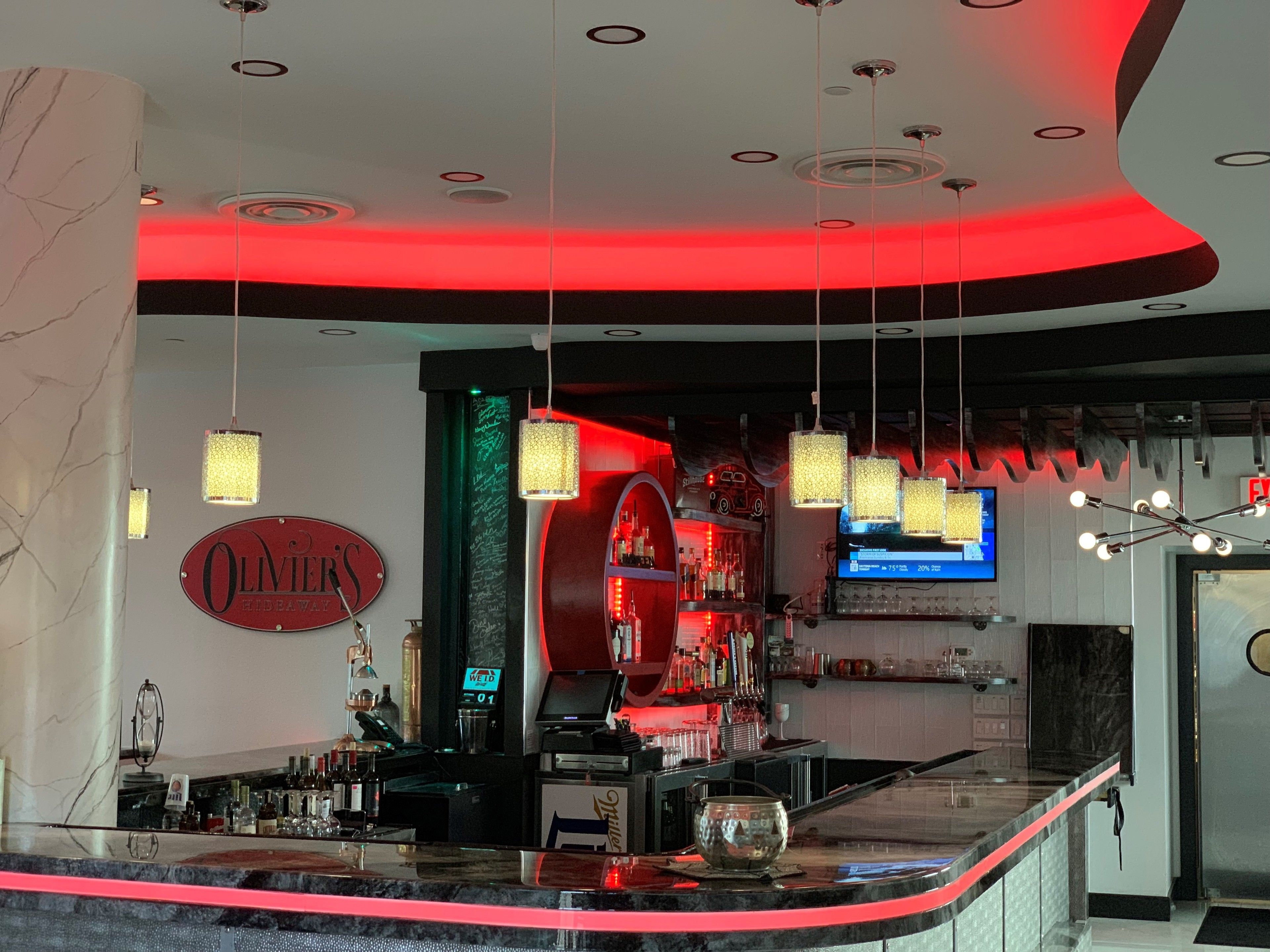Uma área de bar com bancada de mármore, luminárias pendentes e uma tela de televisão exibindo um cardápio.