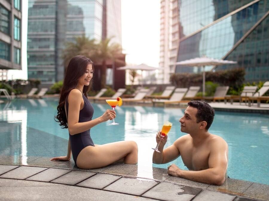 Um casal junto a uma piscina com bebidas, rodeado por edifícios modernos.