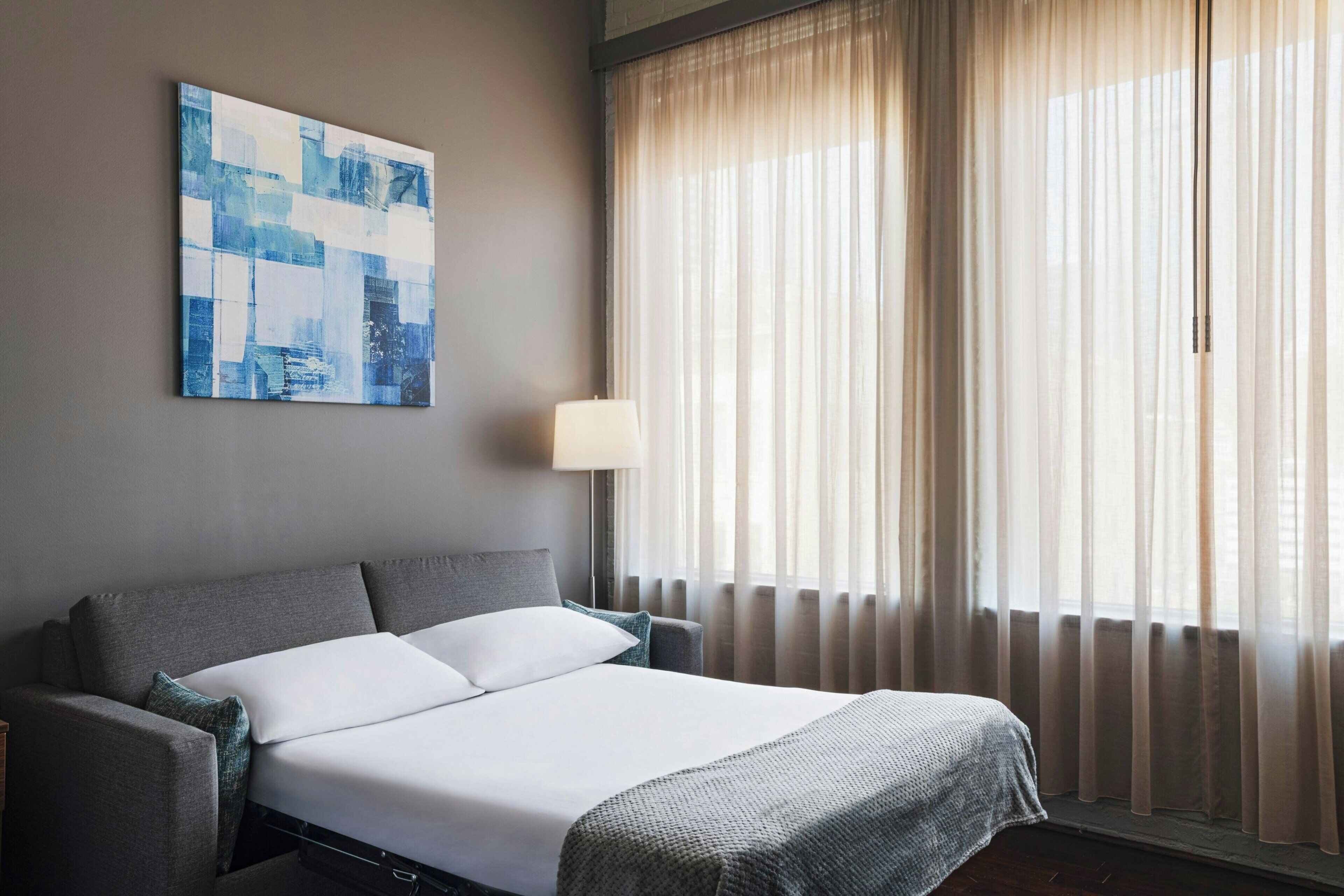 Quarto de hotel com uma cama, um sofá cinzento, uma luminária branca e uma janela com cortinas leves.