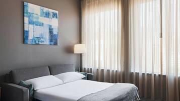 Quarto de hotel com uma cama, um sofá cinzento, uma luminária branca e uma janela com cortinas leves.