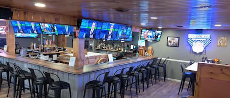 Um bar com várias telas exibindo eventos esportivos, um balcão com utensílios de bar e vários bancos altos pretos.