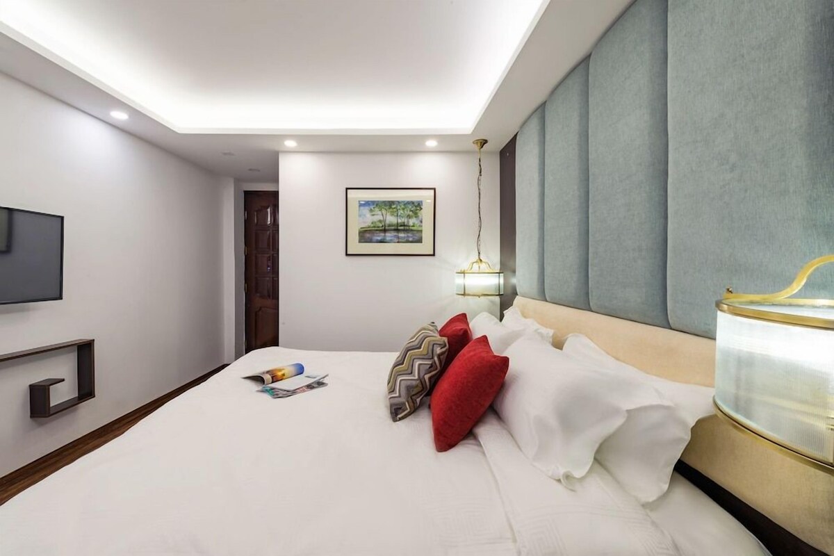 Quarto de hotel moderno com uma cama grande, televisão, um quadro na parede e uma luminária de cabeceira.