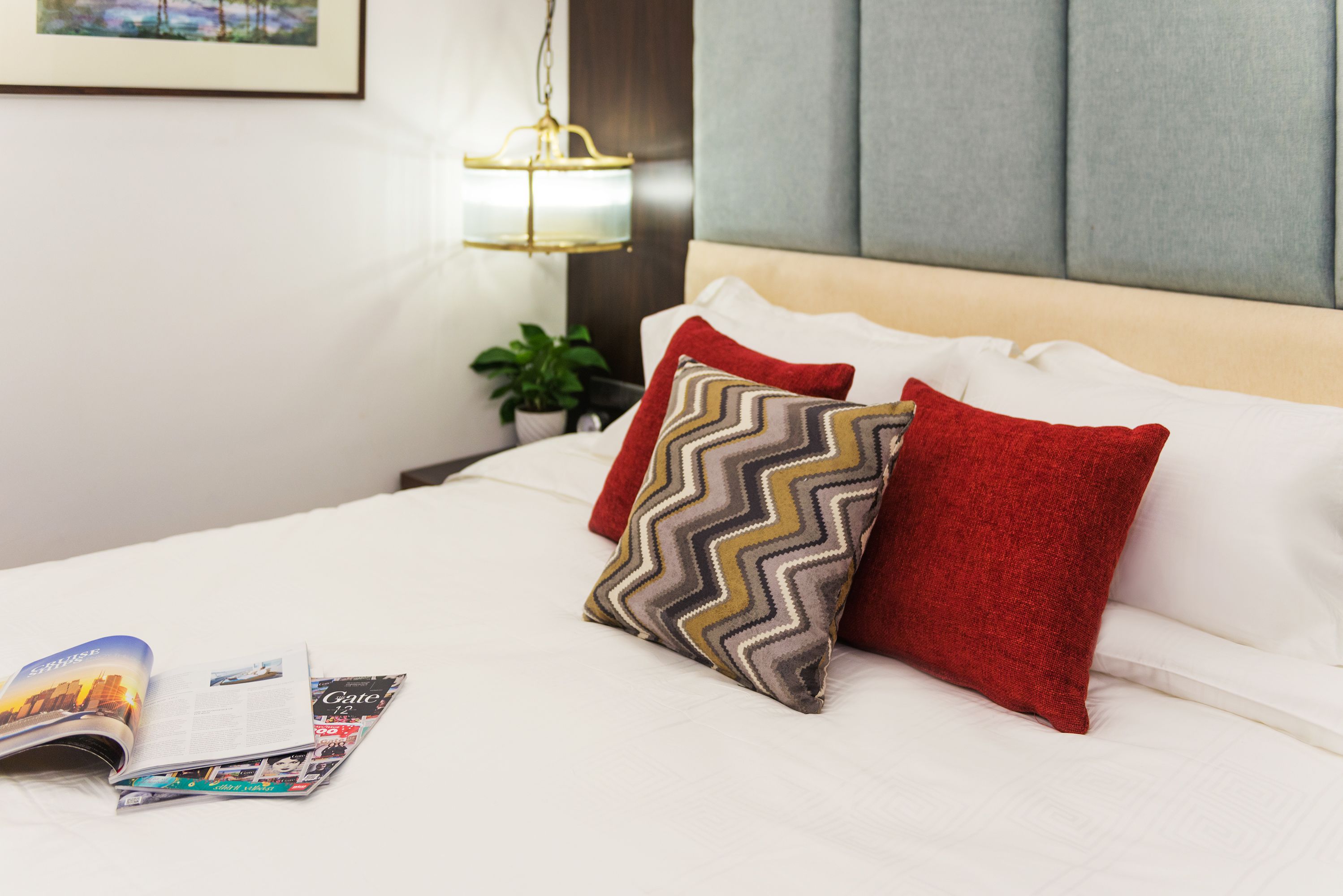 Cama bem arrumada com lençóis brancos, duas almofadas decorativas, uma revista e um abajur.
