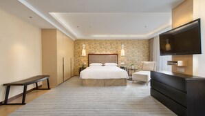 Quarto de hotel com uma cama grande, uma escrivaninha, uma cadeira, uma televisão e um banco.