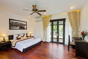 Quarto de hotel com cama, mesas de cabeceira, escrivaninha, cadeira, ventoinha no teto e porta que dá para uma área externa.