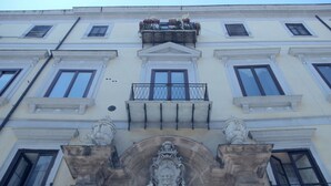 Um edifício com fachada clássica, apresentando um frontão central com relevo escultórico, janelas simétricas e uma varanda com grades.