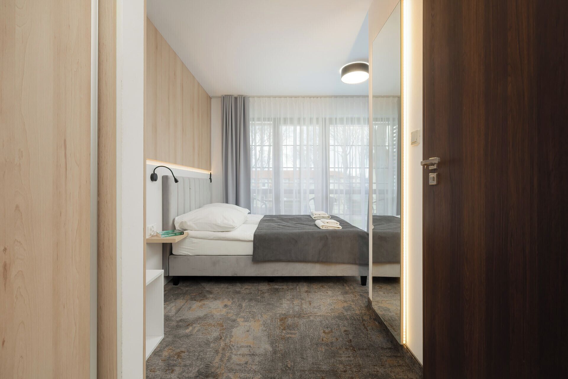 Quarto com cama, mesa de cabeceira, abajur e vista para o exterior pela janela.
