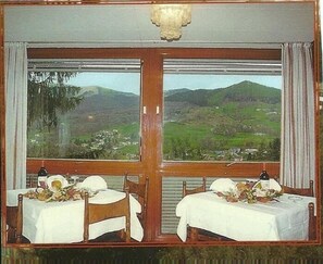 Uma área de refeições com duas mesas preparadas para dois, cada uma com toalha branca e centros de mesa florais. Grandes janelas oferecem vista para uma paisagem montanhosa.