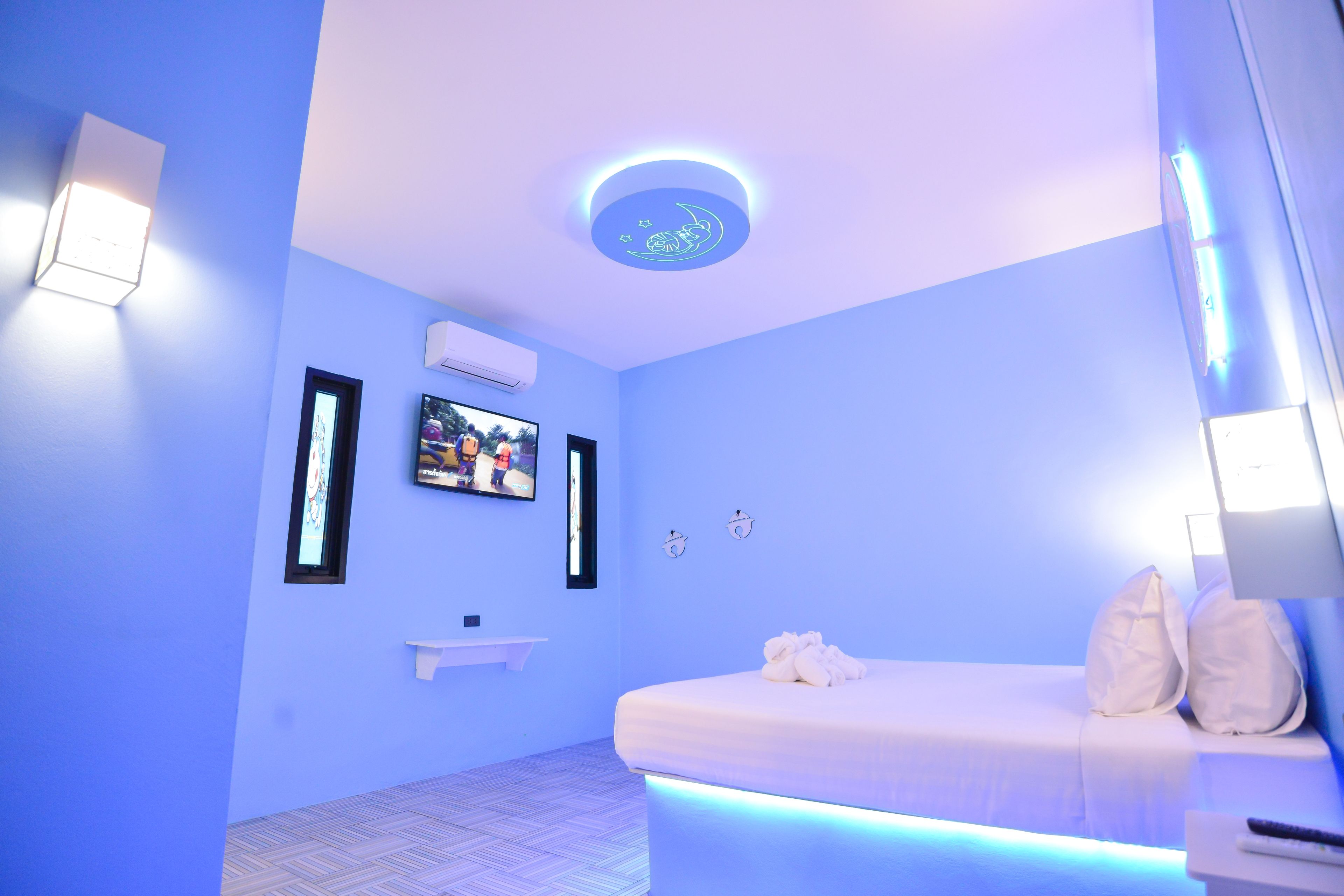 Quarto moderno com parede azul, cama com roupa de cama branca, prateleira pequena branca e luminária no teto.