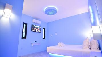 Quarto moderno com parede azul, cama com roupa de cama branca, prateleira pequena branca e luminária no teto.