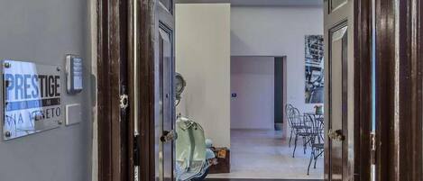 Entrada de hotel com um letreiro indicando quartos 'Prestige' e 'Veneto'. Há uma área de estar com cadeiras e uma mesa ao fundo.