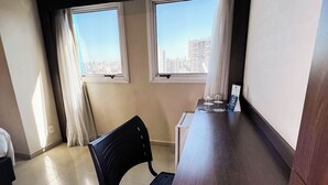 Quarto com escrivaninha de madeira, duas janelas com cortinas, ar condicionado e uma cadeira.