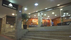 Entrada de hotel com uma placa indicando "Royal Center Hotel", uma escadaria que leva ao saguão e uma planta em vaso à esquerda.