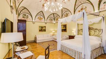 Quarto com cama de dossel, área de estar com sofá e poltrona, escrivaninha com luminária e lustre.