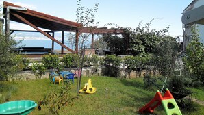 Uma área externa gramada com uma estrutura de madeira, vista para o mar e diversos equipamentos de recreação infantil.