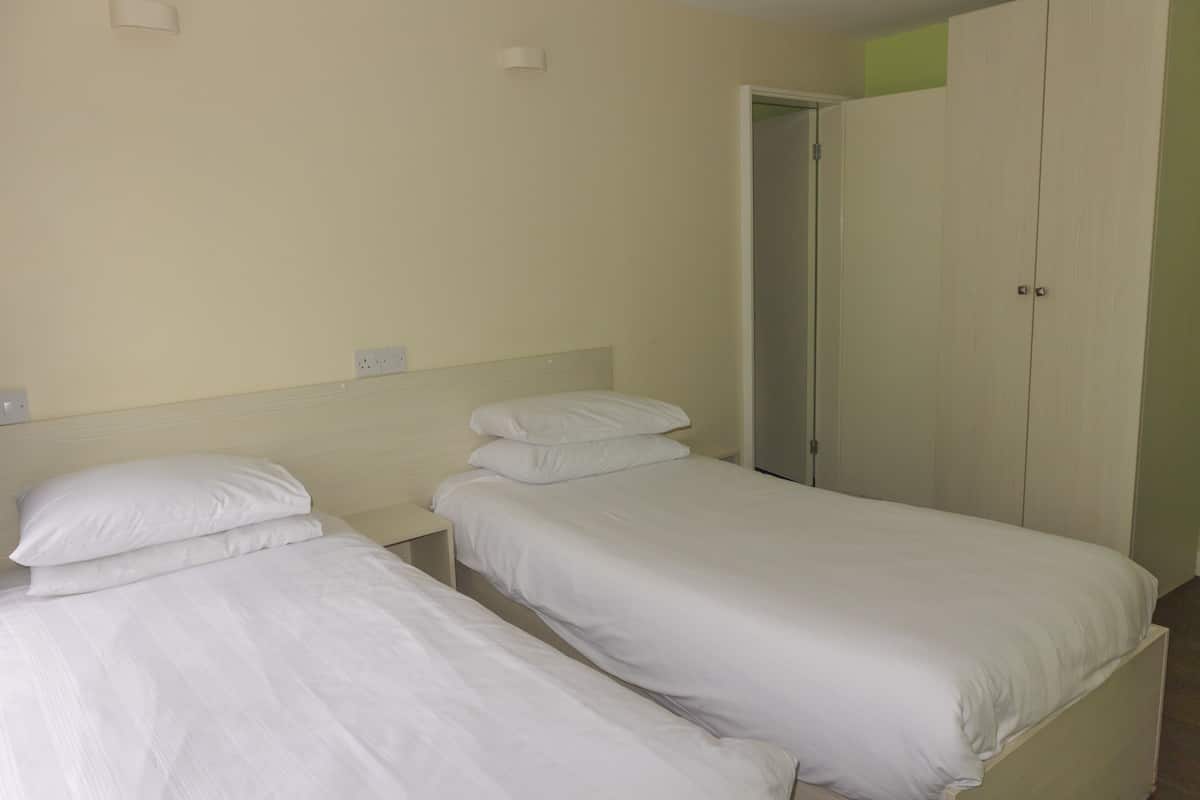 Quarto com duas camas de solteiro, um guarda-roupa e uma porta que dá para outro cômodo.