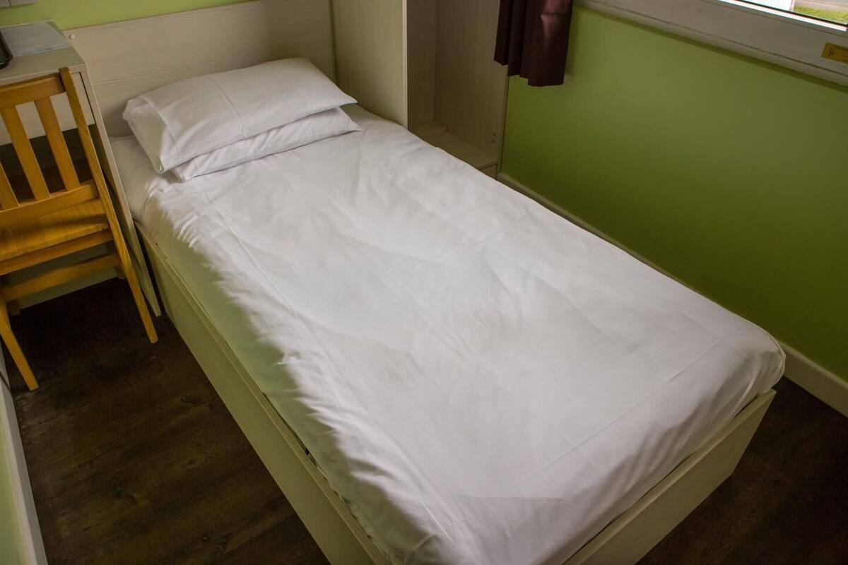 Uma cama de solteiro com roupa de cama branca num quarto com paredes verdes e soalho de madeira.